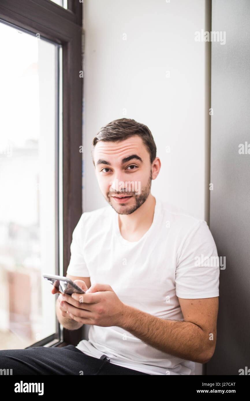 White man sitting on a windowsill in a home environment holding a phone ...