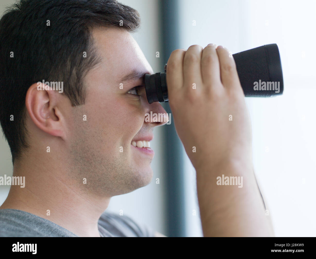 Young man standing looking through a glass window with binoculars as he ...