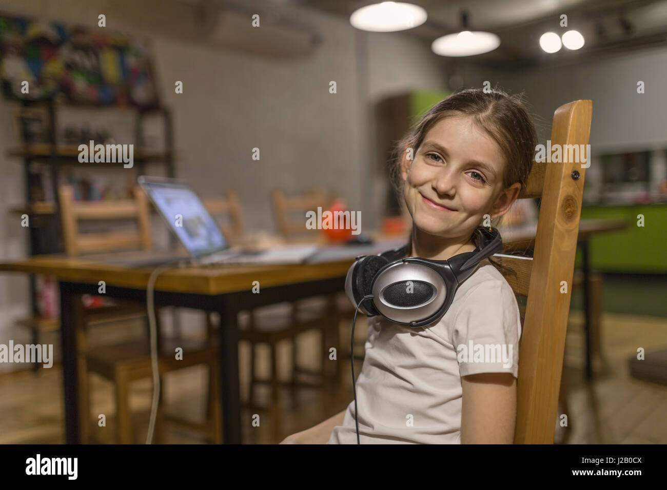 Portrait of smiling girl wearing headphones around neck while sitting on wooden chair at home ...