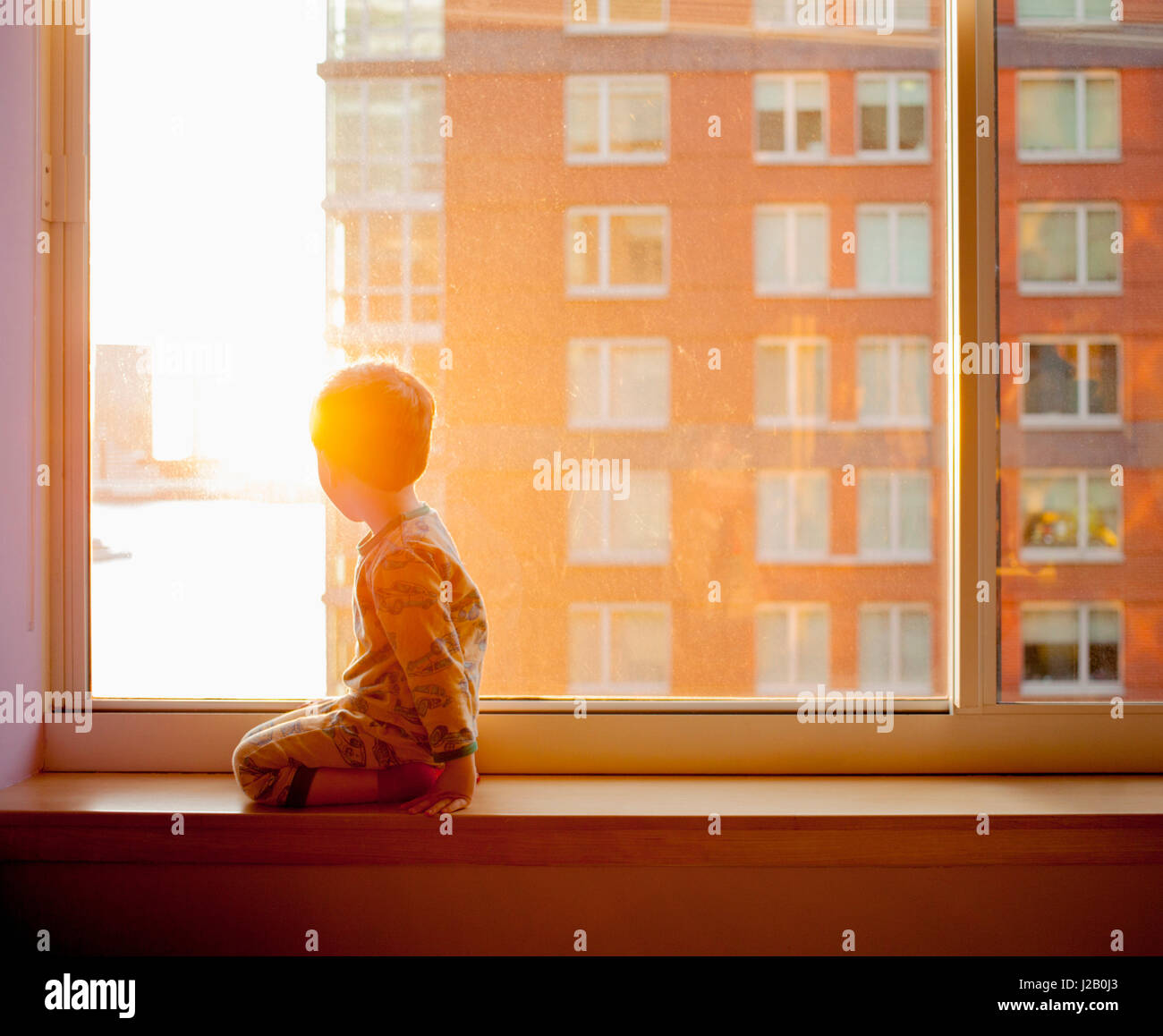 Side view of boy looking out while kneeling on window sill at home ...