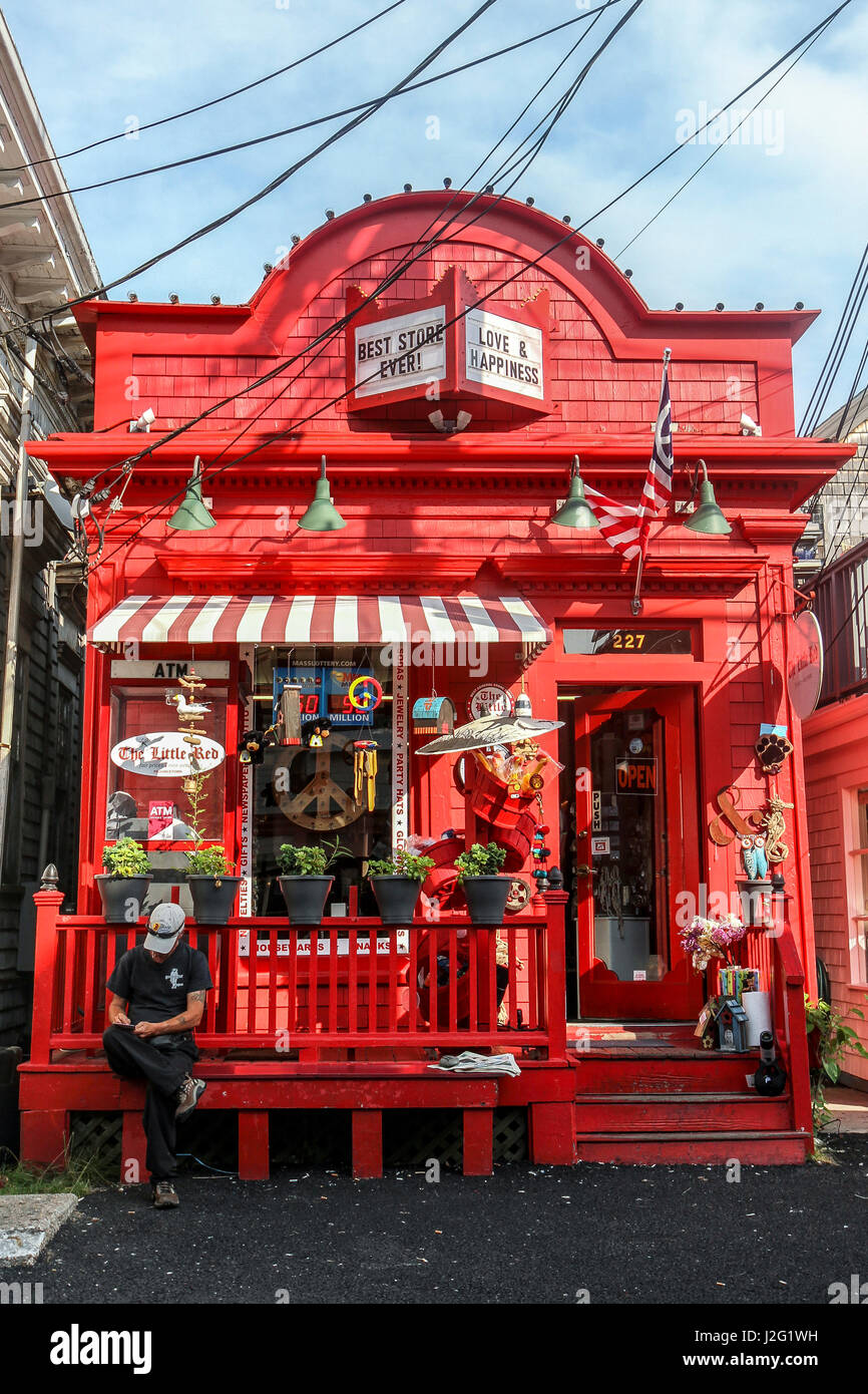 A man sits in front of a bright red building housing a shop that ...