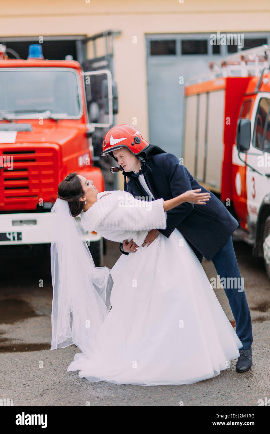 Firesman holding on hands his new wife with fire trucks at background ...