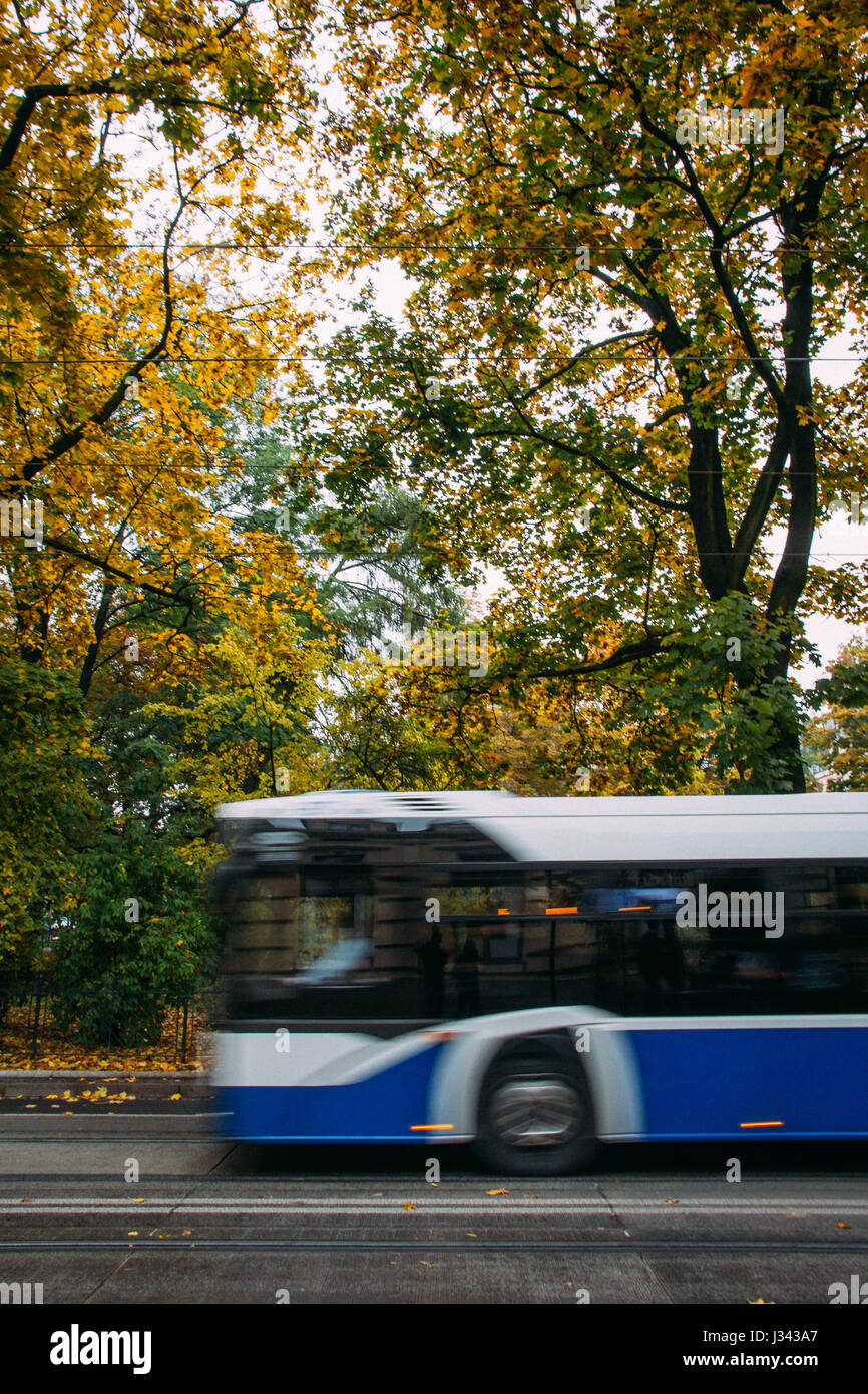 A blue public bus moves across the frame down the street with trees and ...