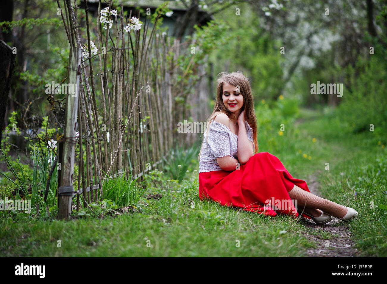 Portrait of sitting beautiful girl with red lips at spring blossom garden on green grass, wear ...