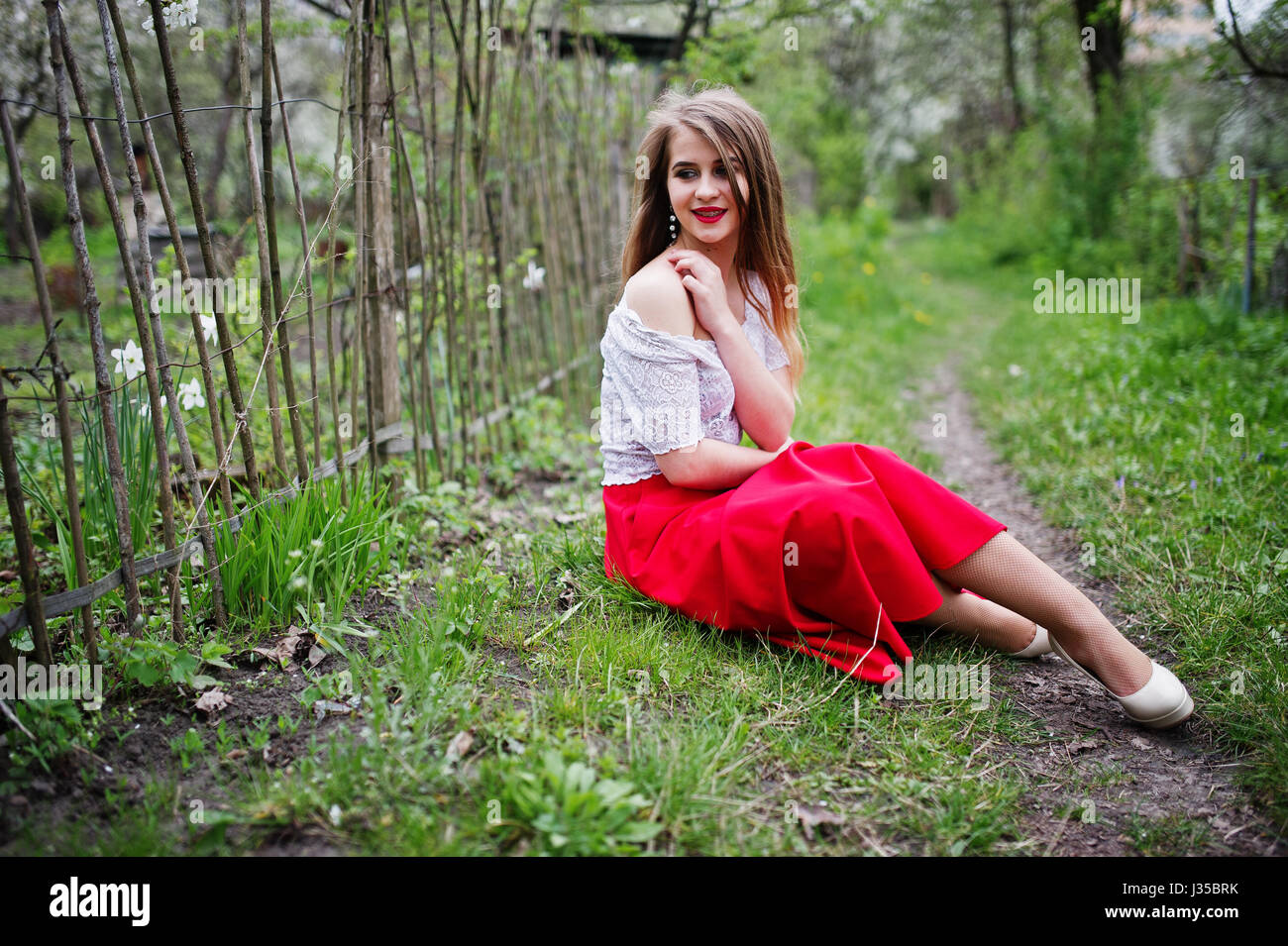 Portrait of sitting beautiful girl with red lips at spring blossom garden on green grass, wear ...