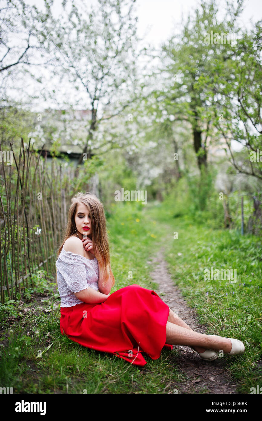 Portrait of sitting beautiful girl with red lips at spring blossom garden on green grass, wear ...