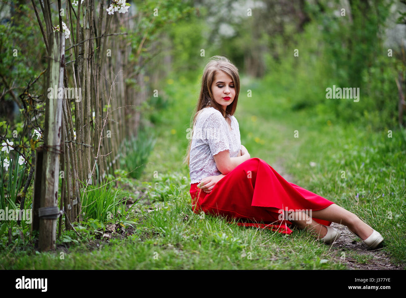 Portrait of sitting beautiful girl with red lips at spring blossom garden on green grass, wear ...
