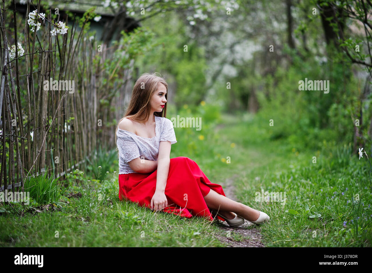 Portrait of sitting beautiful girl with red lips at spring blossom garden on green grass, wear ...
