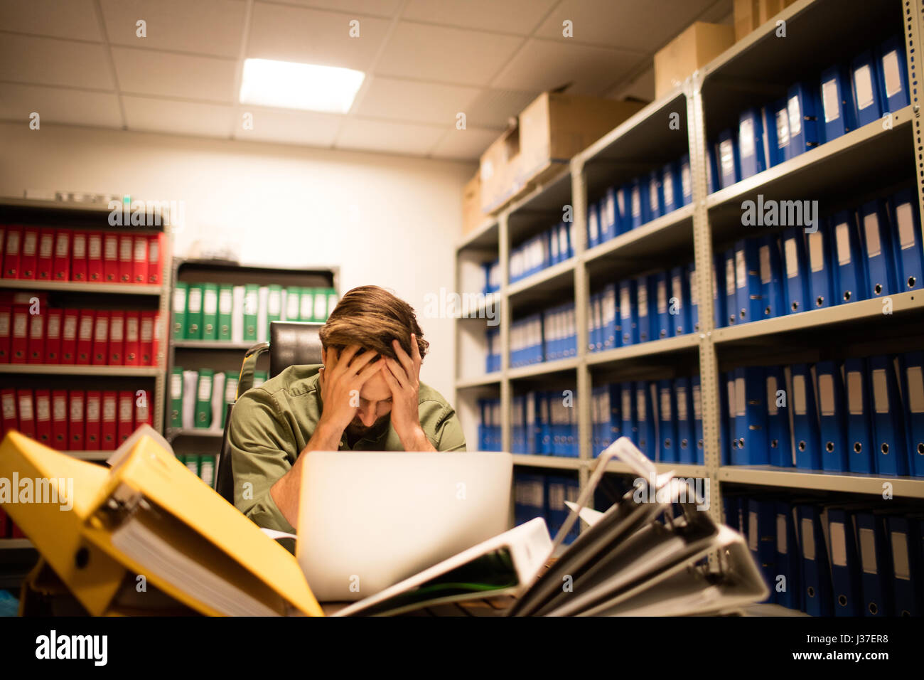 Frustrated businessman with pile of files and laptop in storage room ...