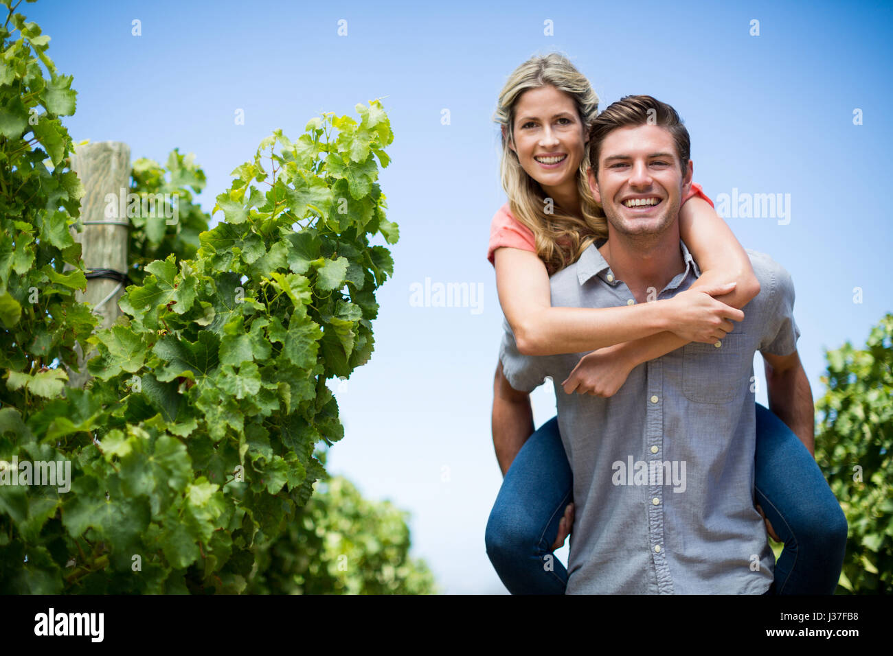 Portrait of happy young couple piggybacking at vineyard against blue ...
