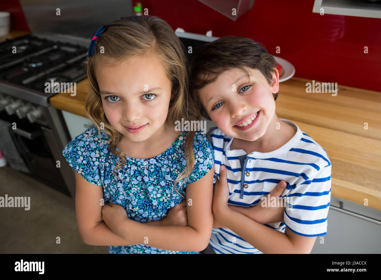 Portrait of happy siblings standing with arms crossed in kitchen at ...