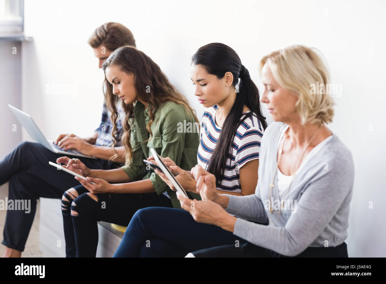 Business people using technologies while sitting on seat against wall ...