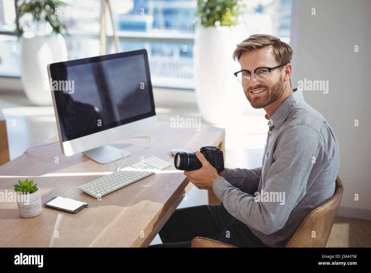 Portrait of smiling executive holding digital camera at desk in office ...