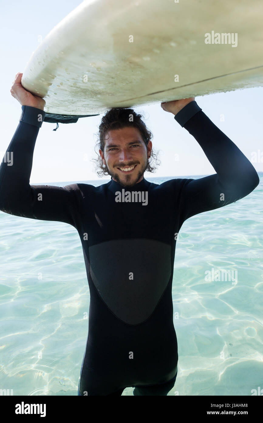 Portrait of smiling surfer in wetsuit carrying surfboard over head at ...