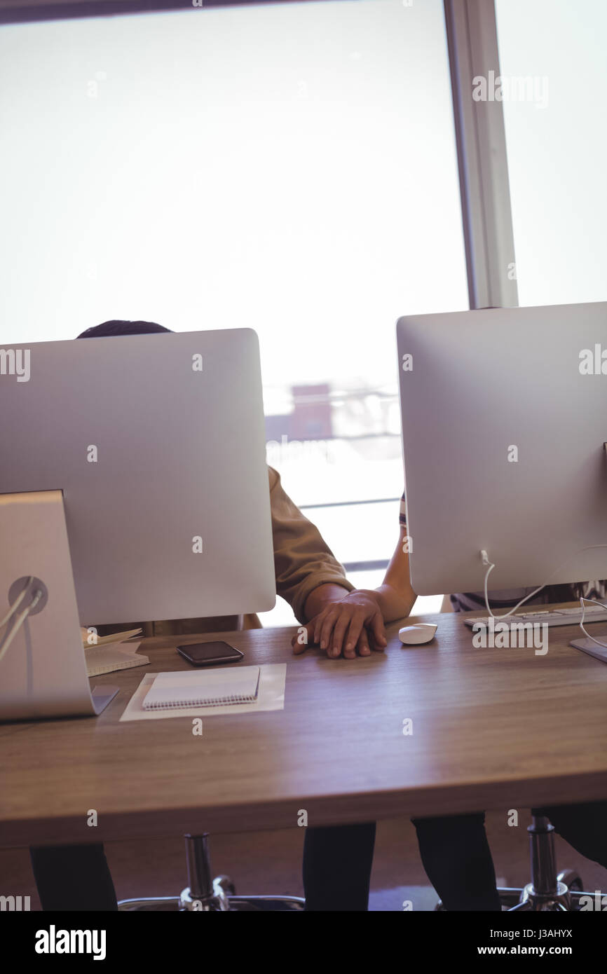 Business couple holding hands while sitting behind computers in office ...