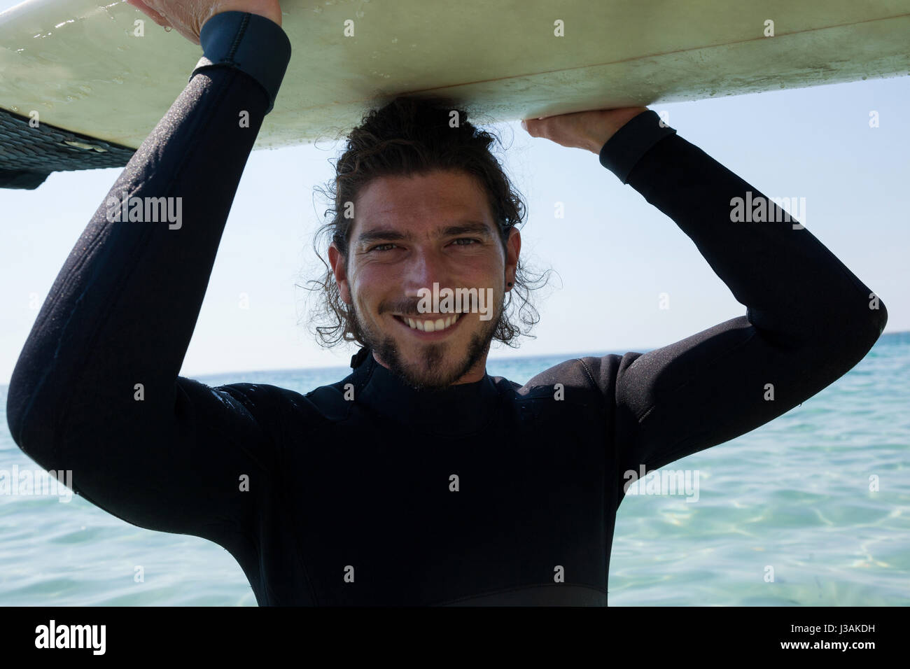 Portrait of smiling surfer in wetsuit carrying surfboard over head at ...