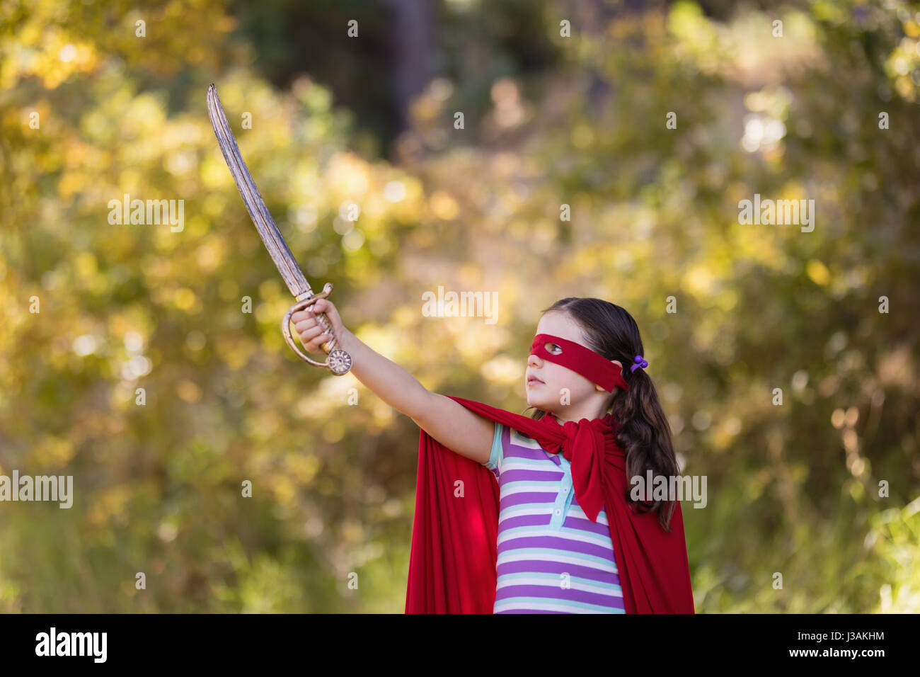Little girl holding sword while wearing superhero costume in forest ...