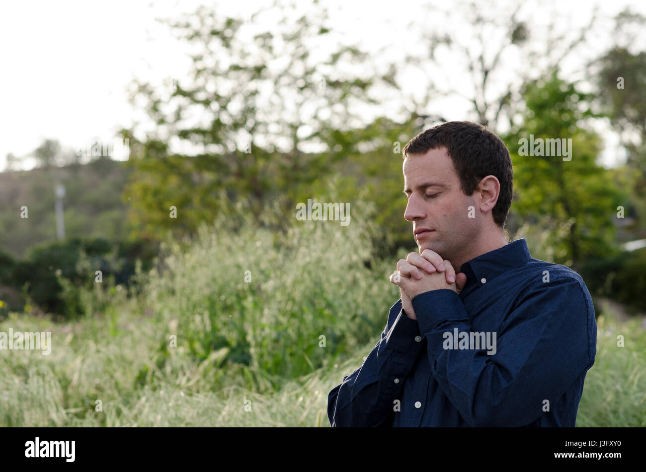 Man praying alone outside in a grassy field with hands folded and eyes ...