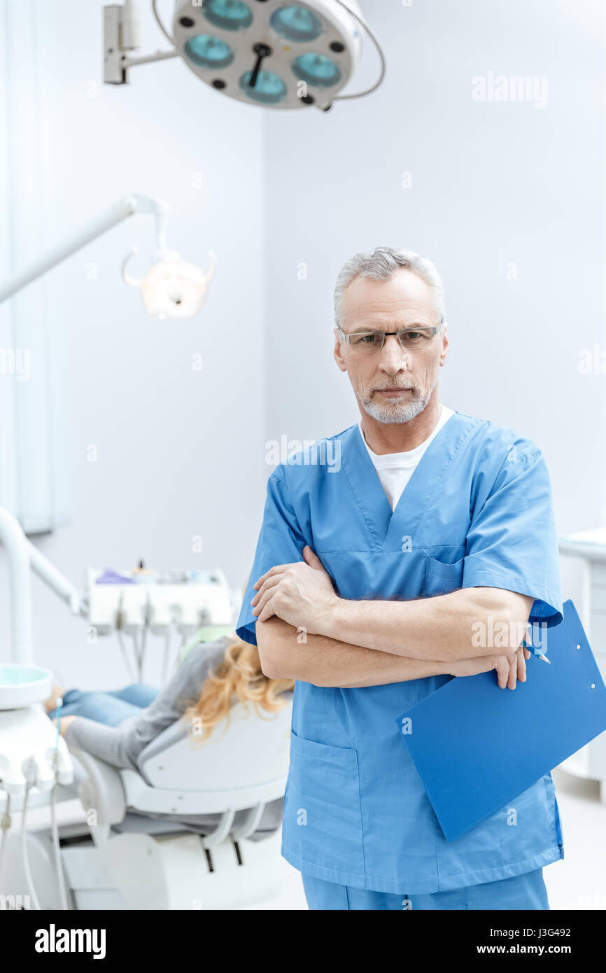 senior dentist in uniform with clipboard in dental clinic with patient ...