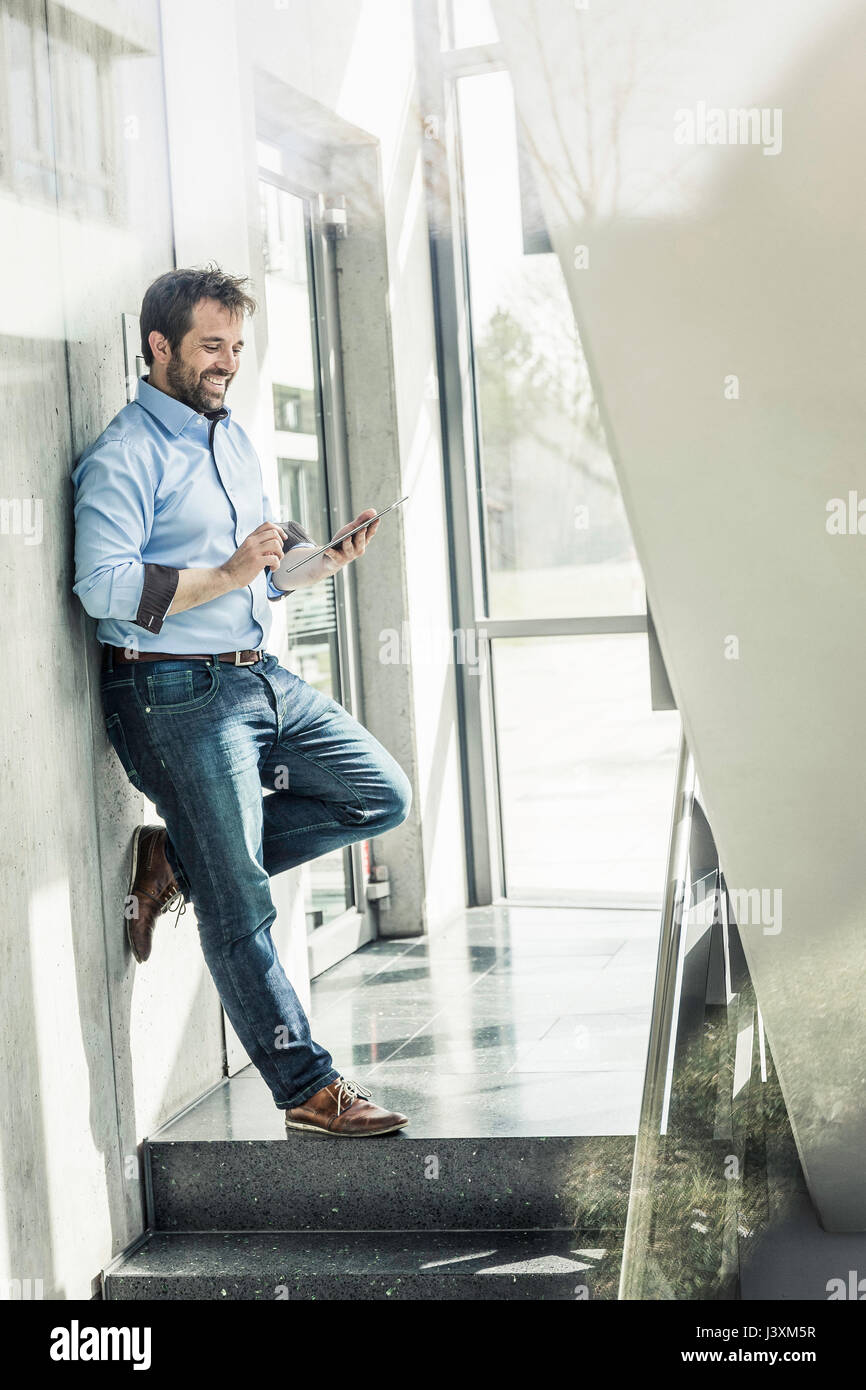 Businessman leaning against office wall using digital tablet ...