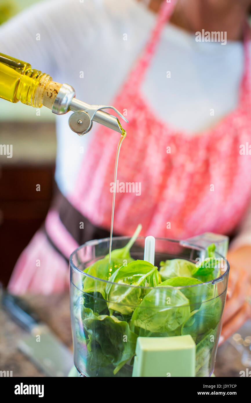 Mature woman pouring olive oil into food processor containing basil ...