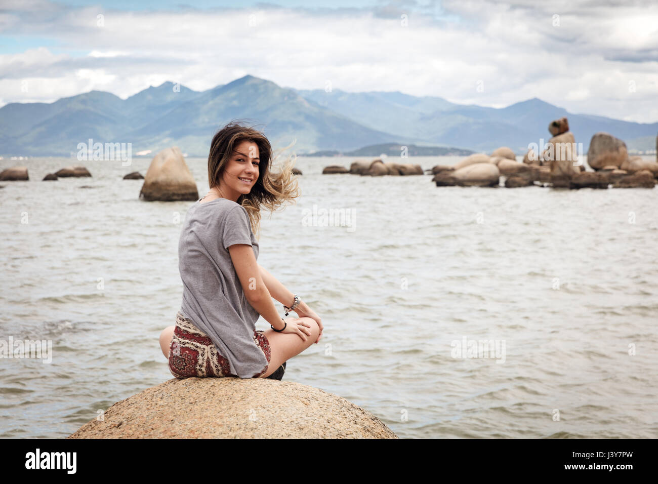 Rear view of teenage girl sitting on rocks looking over shoulder at ...