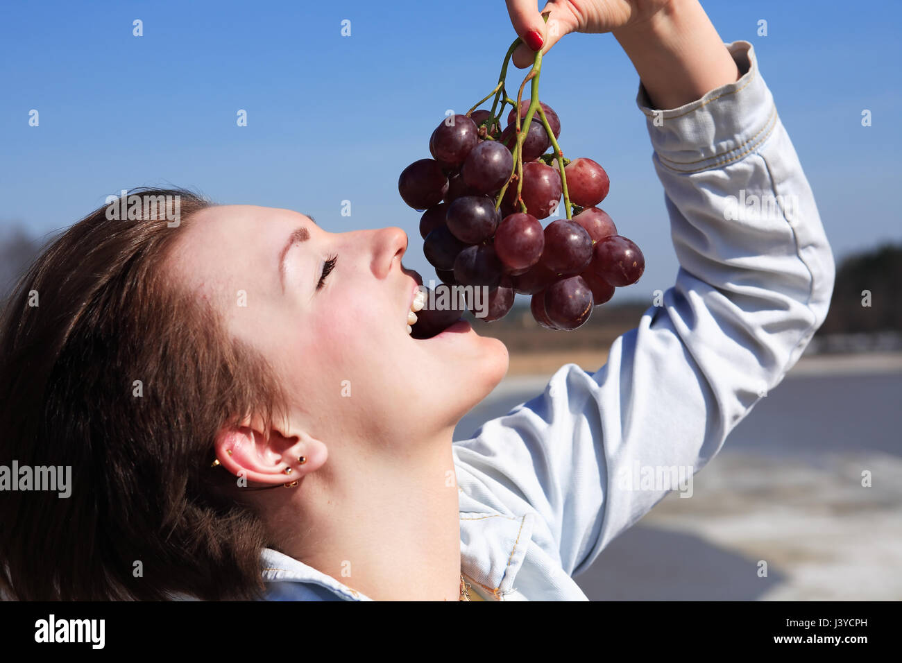 Beautiful young girl eating red grapes outdoor on blue sky background ...