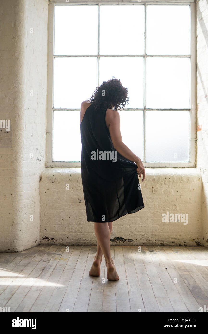 rear view of a young woman wearing a black dress standing by the window ...