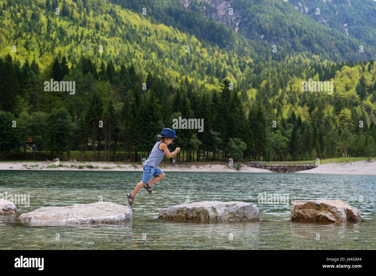 Adorable young boy with a hat crossing river or water jumping from rock ...