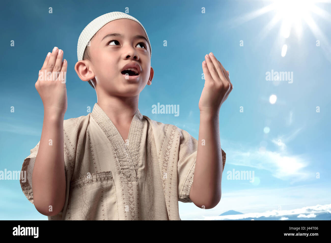 Cheerful asian muslim child in traditional dress praying with blue sky ...