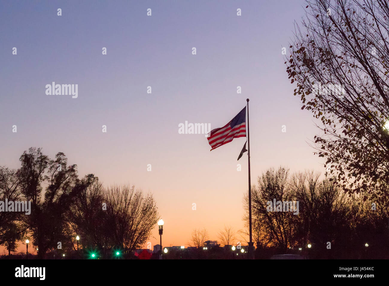 American flag with trees around and city lights at sunset Stock Photo ...