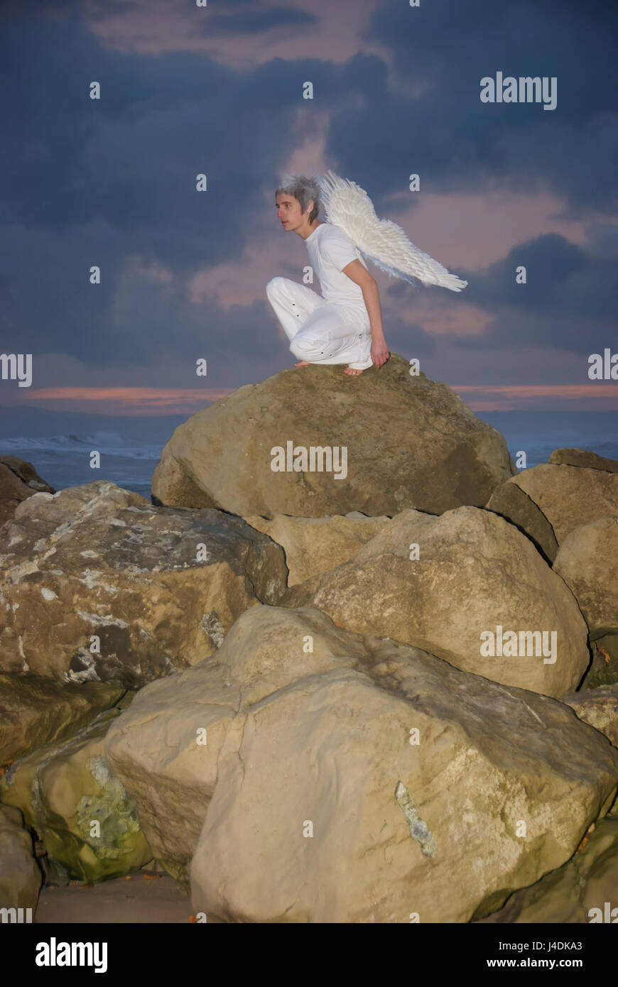Young man dressed in white with angel wings on the rocks by the sea in ...
