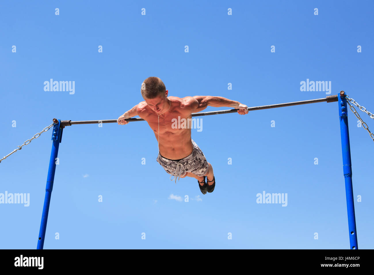 Young strong athlete doing exercise on horizontal bar against blue sky ...