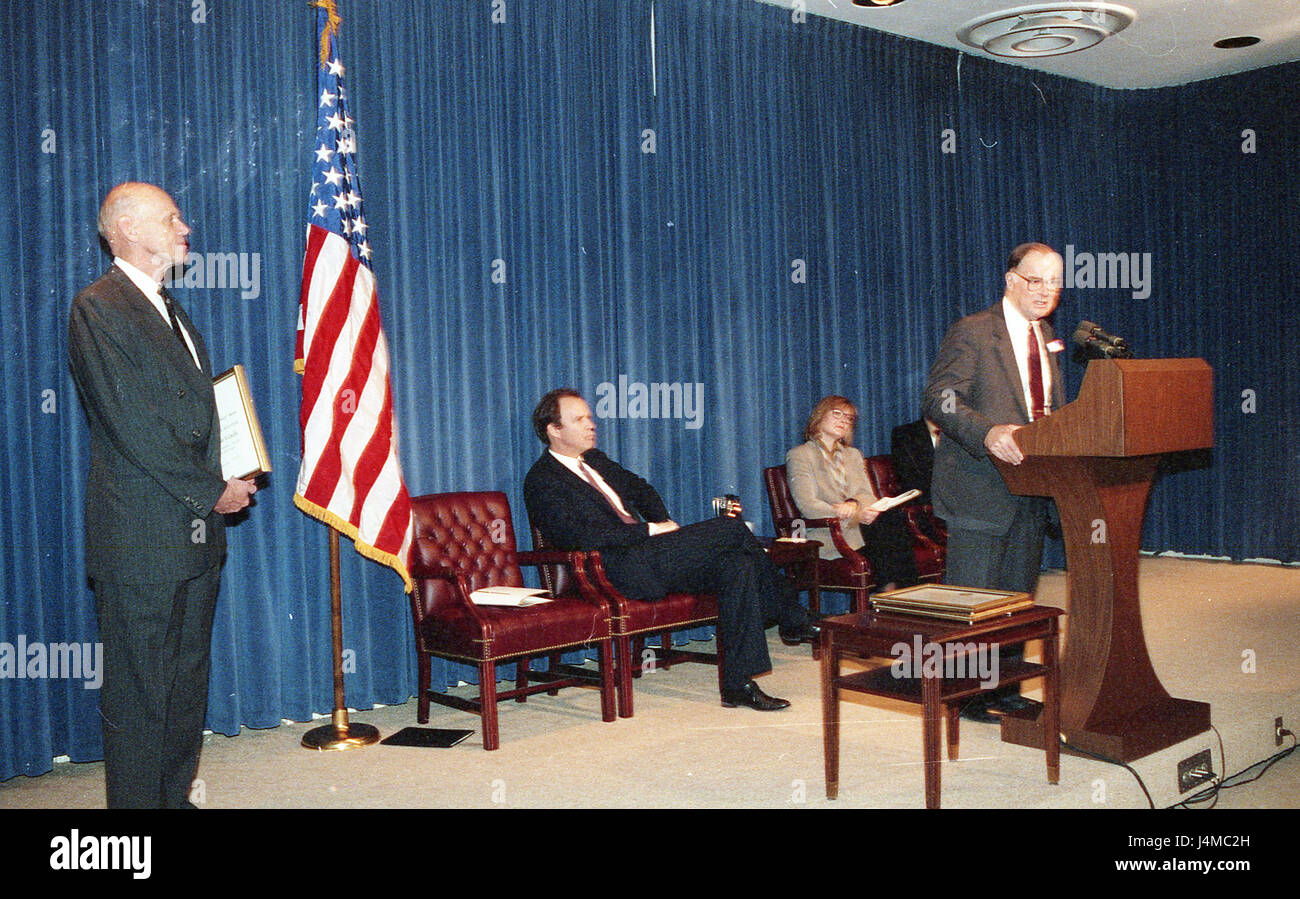 A MAN AT THE PODIUM GIVING A SPEECH WITH THREE OTHER PEOPLE WATCHING ...