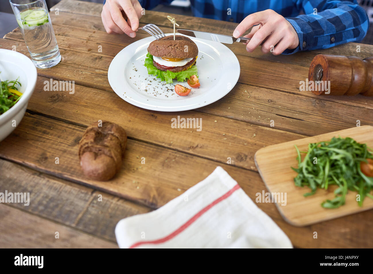 Hungry human with fork and knife eating hamburger by wooden table Stock ...
