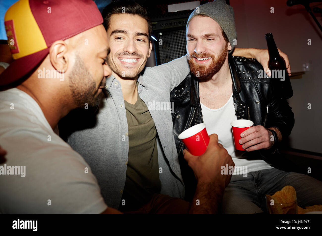 Three smiling young men enjoying party in night club, drinking beer and ...