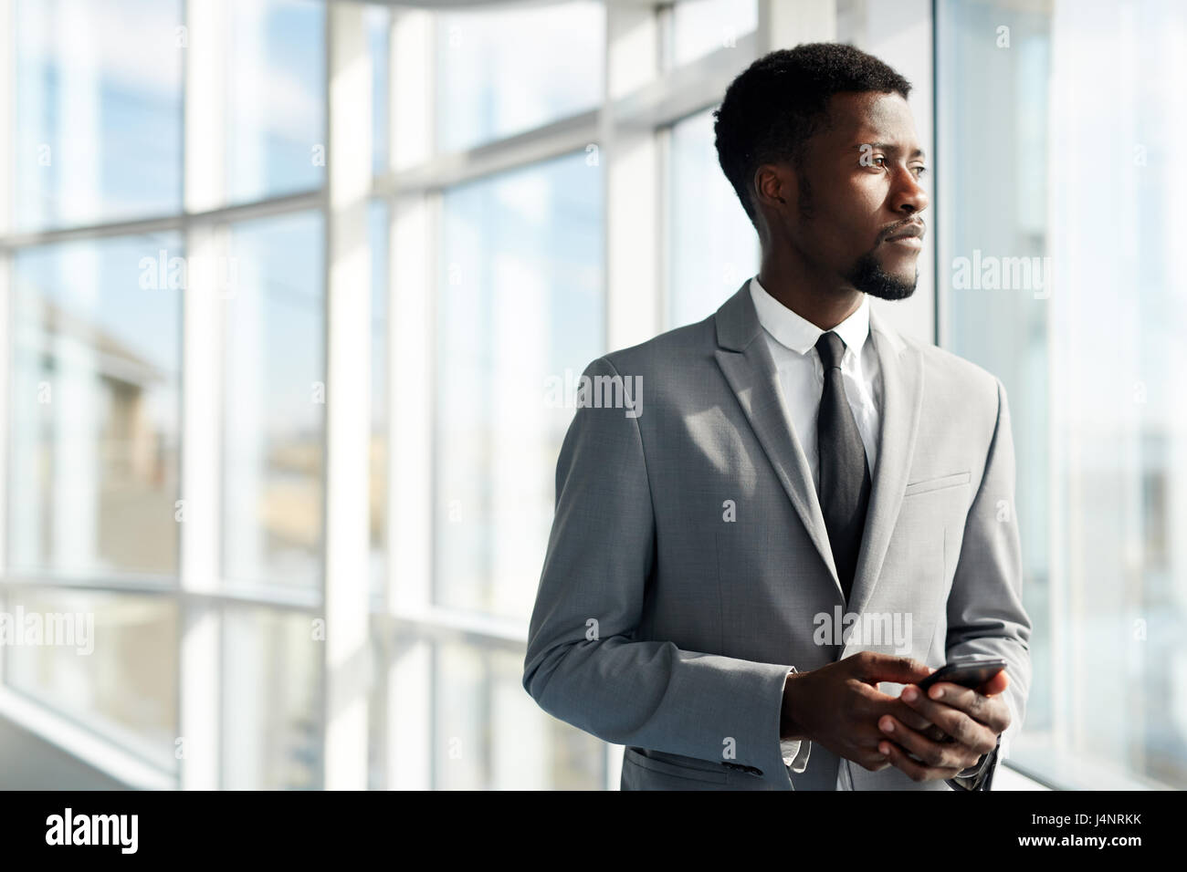 Confident leader with smartphone looking through office window Stock ...