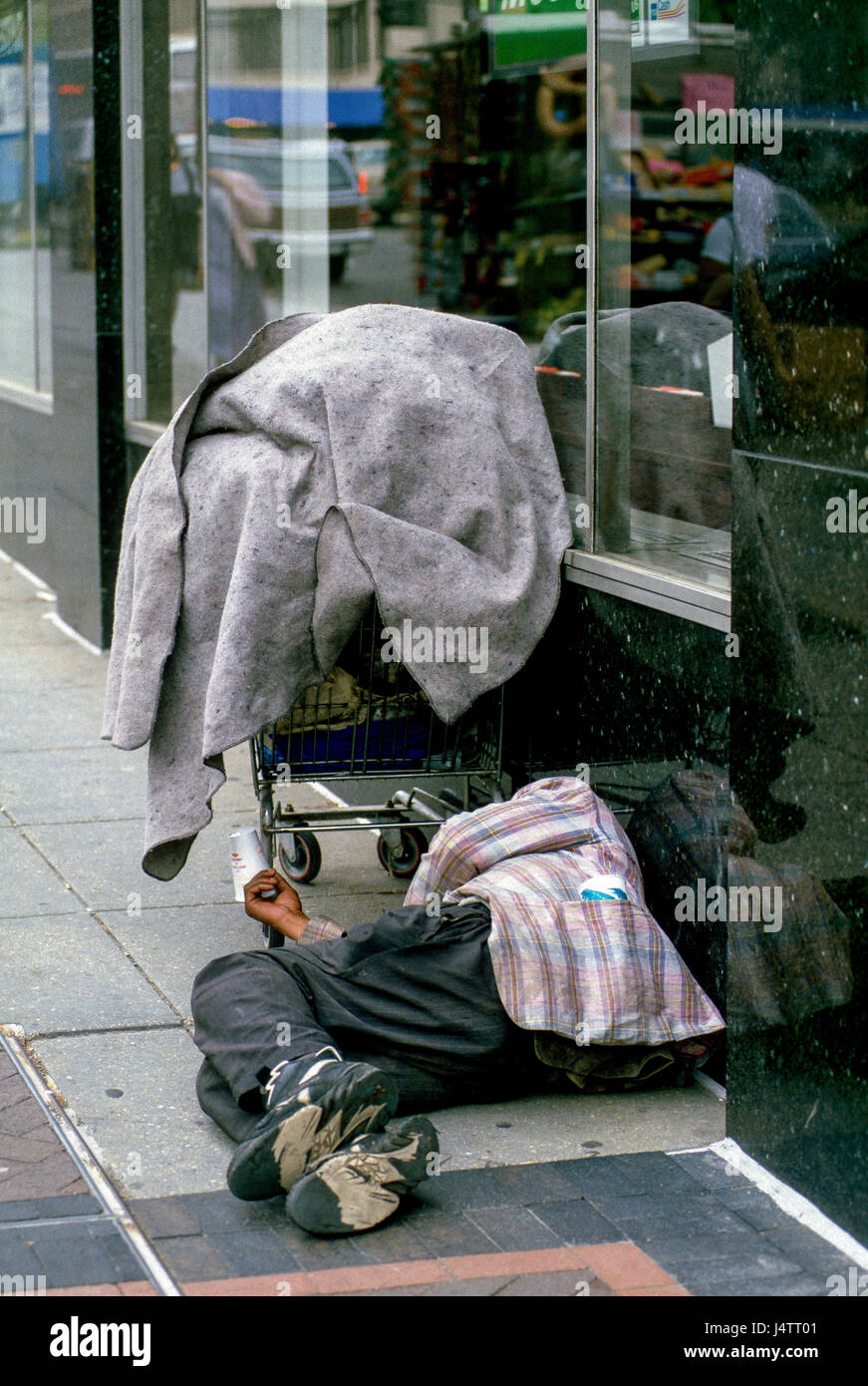 A homeless person lays sleeping on the sidewalk with his grocery cart ...