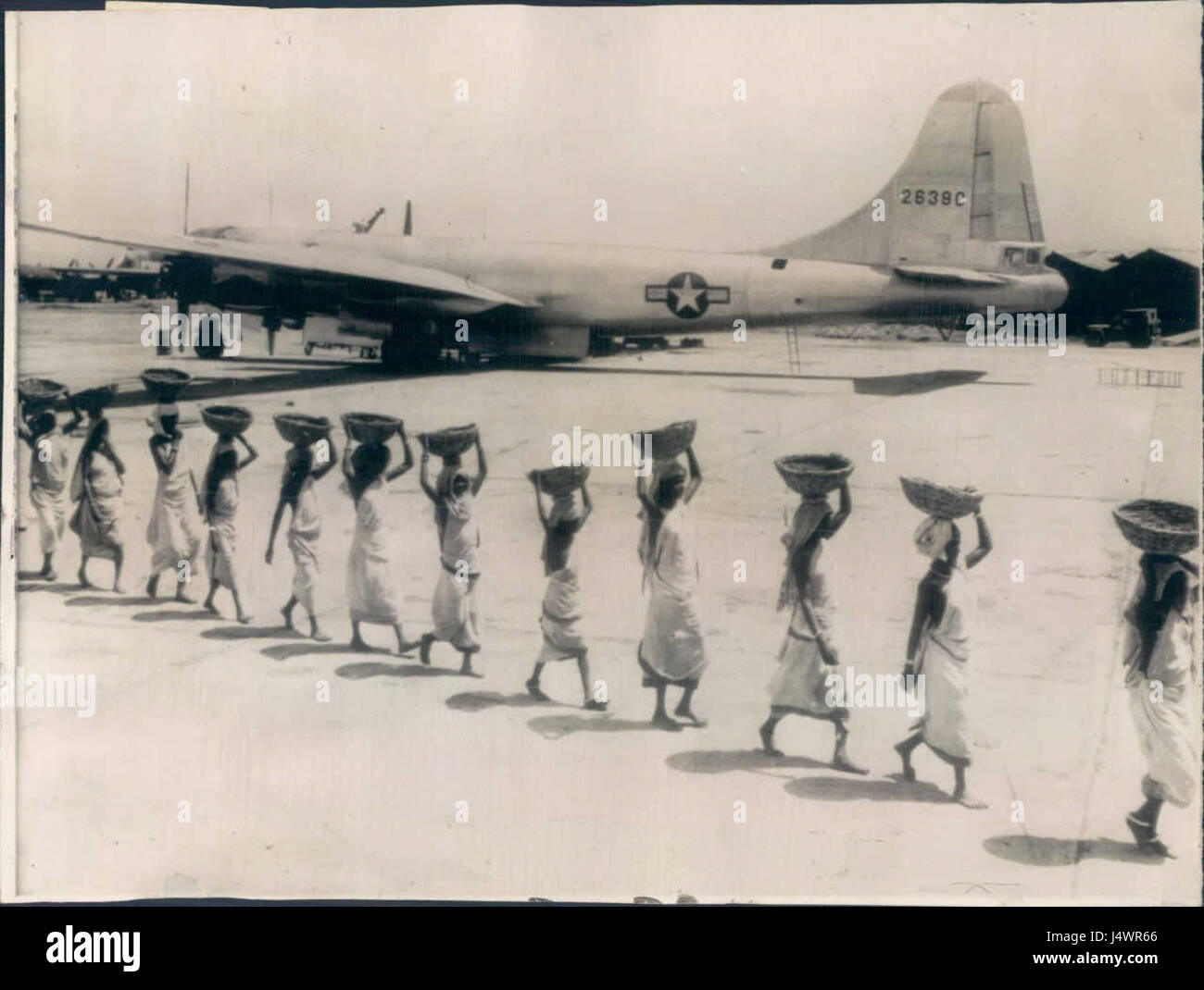 In 1944, women porters in a war-torn area carry head loads past a B-29 ...
