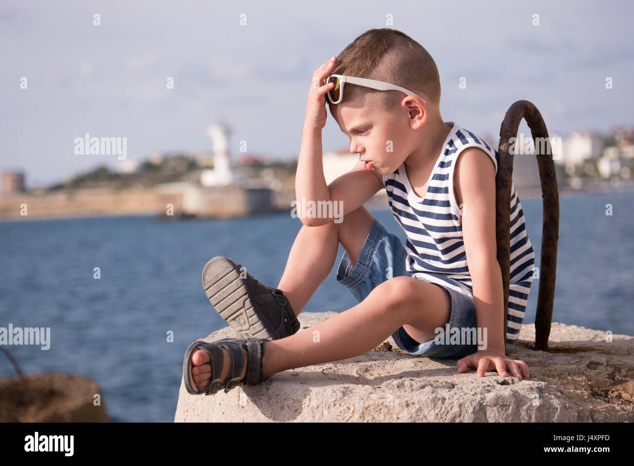 thinking little boy in sunglasses and sailor vest sitting on breakwater ...