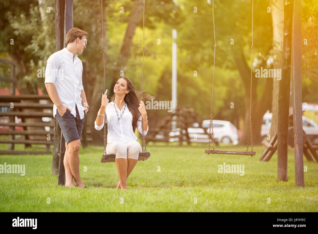 Attractive couple in park on swing, girl sitting on swing man standing ...