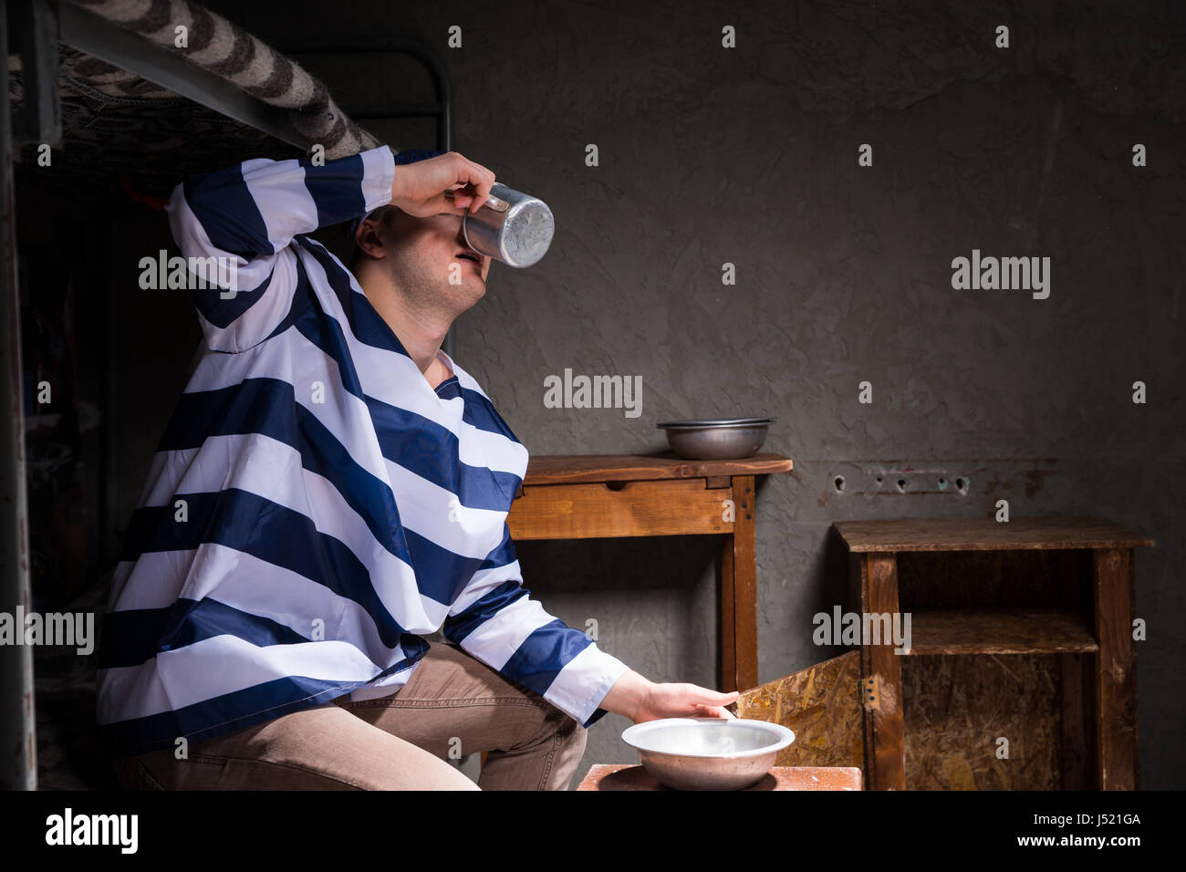 Male prisoner drinking from an aluminum cup sitting on a bed in a small ...