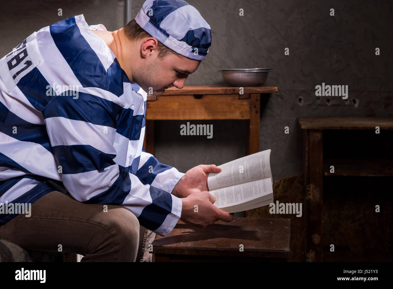 Young male prisoner wearing prison uniform reading a book or a bible ...