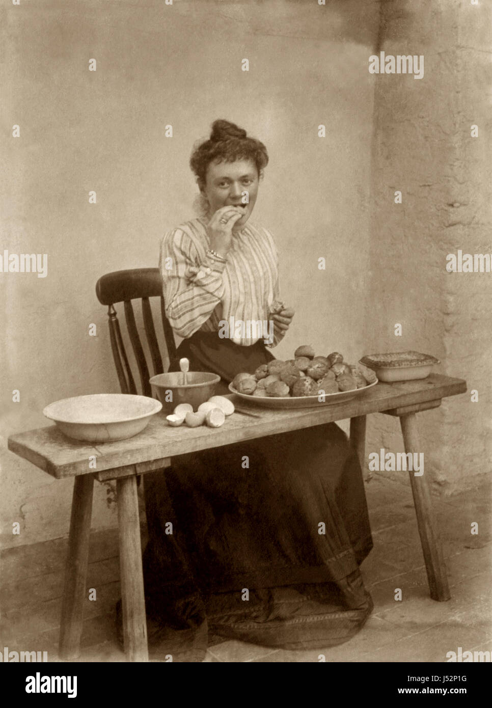 A Victorian woman seated at a trestle table eating cakes and buns ...