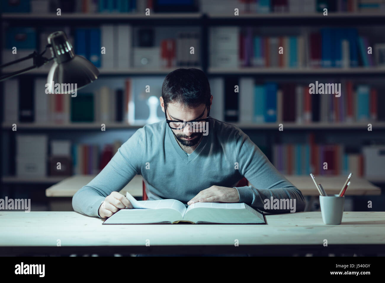 Smart confident young man studying late at night, he is sitting at desk ...