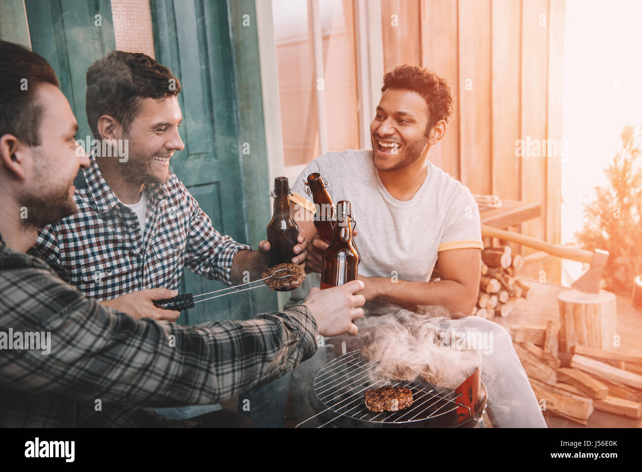 happy young friends making barbecue and drinking beer on porch with ...