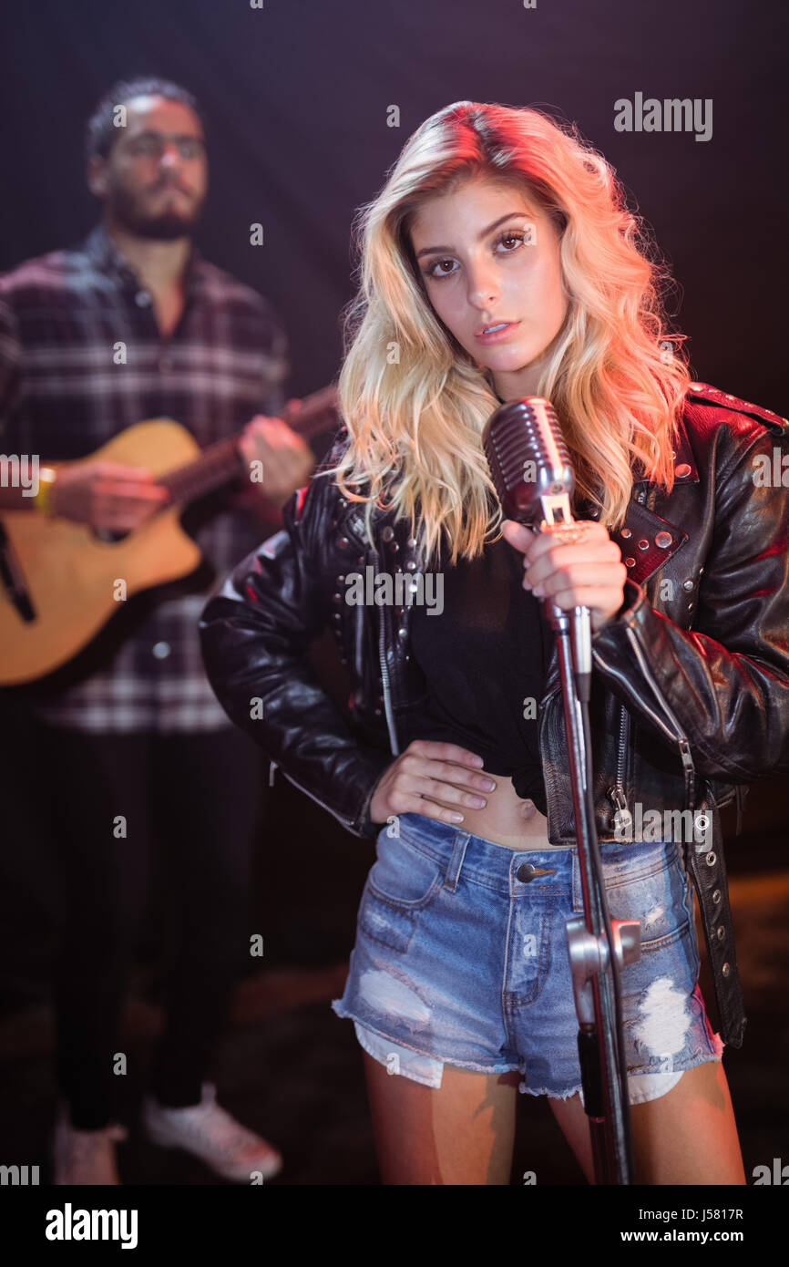 Portrait of young female singer with hand on hip while holding mic at ...