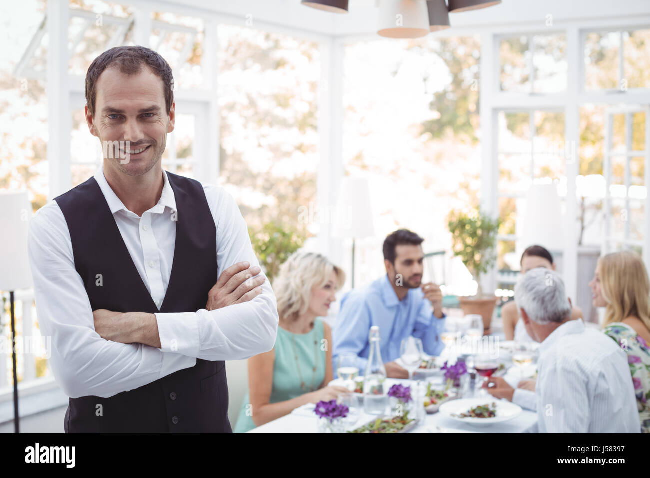 Portrait of smiling waiter standing with arms crossed while friends ...