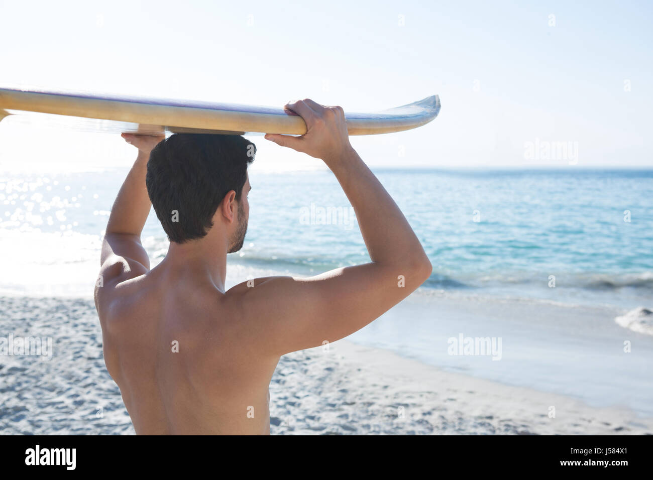 Rear view of shirtless man carrying surfboard at beach on sunny day ...