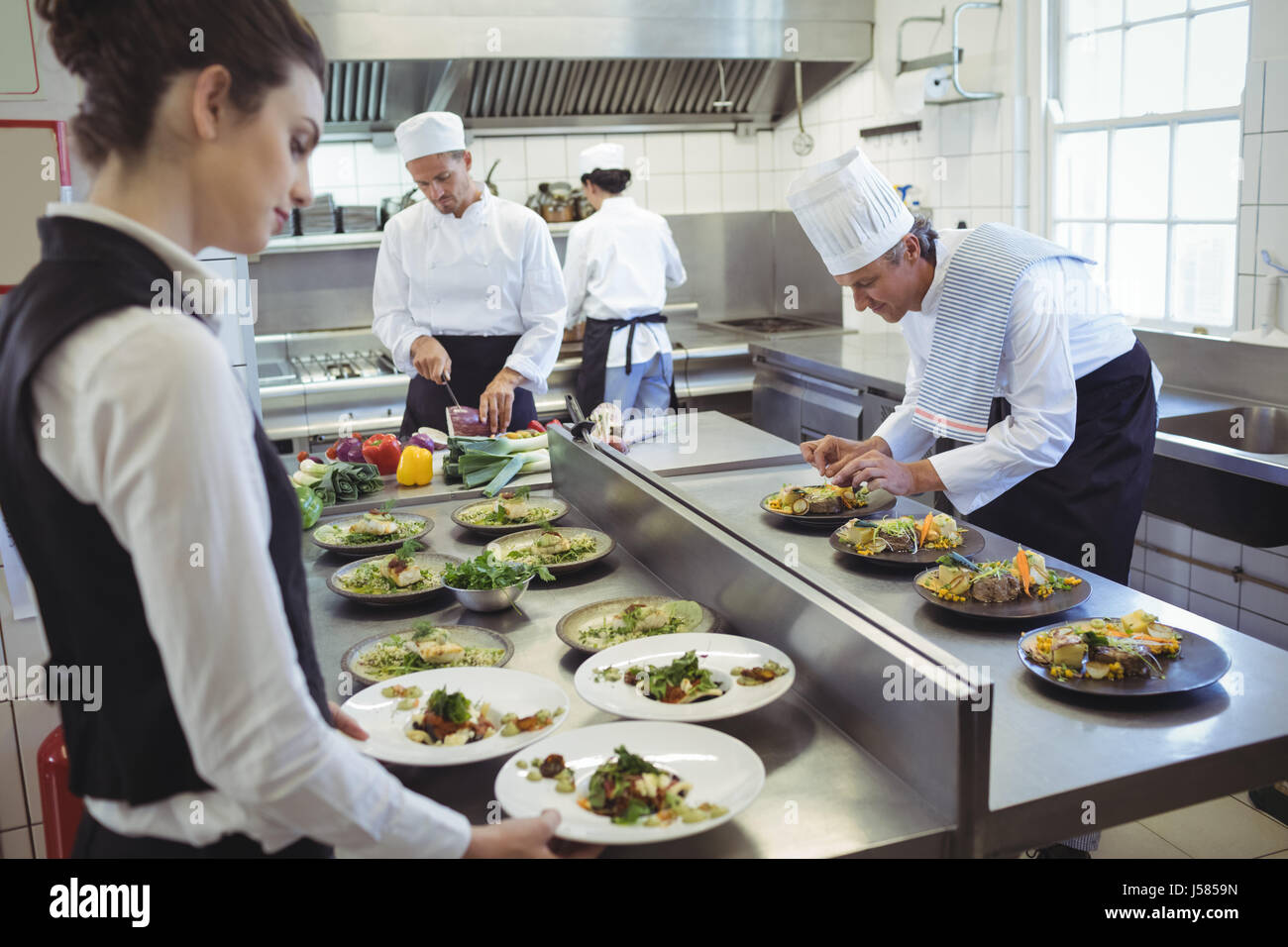 Waitress taking food dishes from order station and chefs preparing food ...
